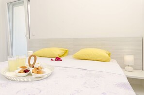 Quarto de hotel com uma cama, uma mesa de cabeceira com abajur, um serviço de jantar com alimentos e bebidas, e uma janela com vista.