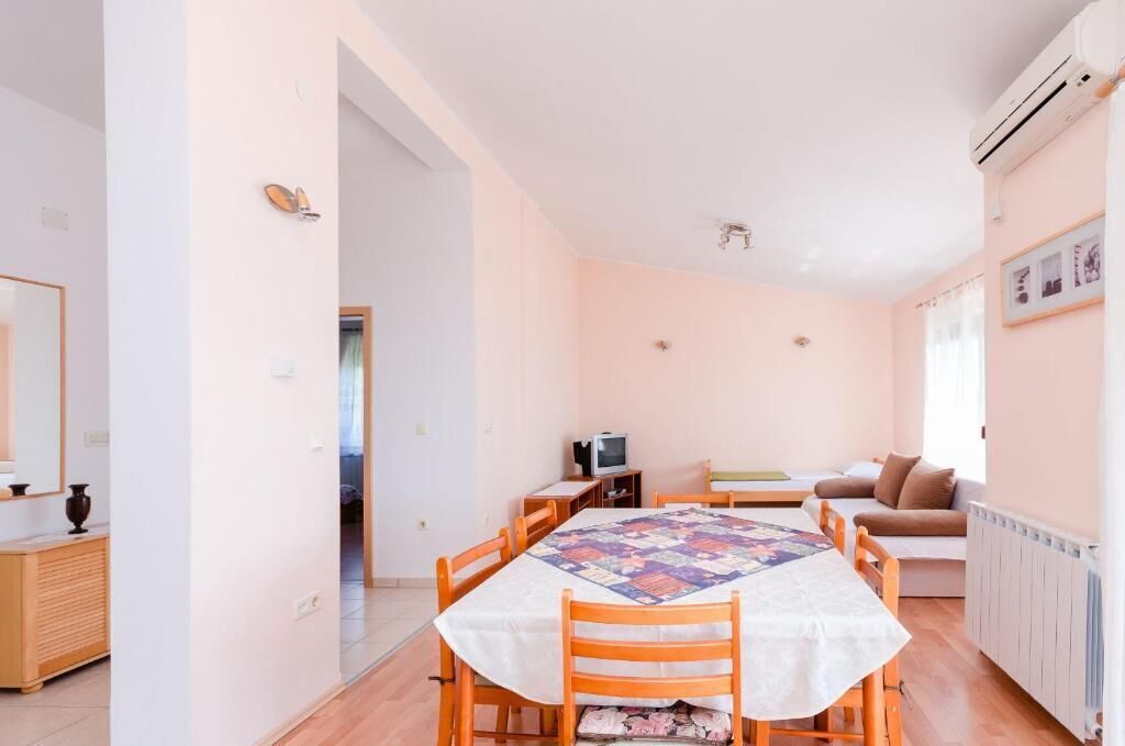 Uma sala de jantar com uma mesa coberta por um colcha de patchwork, cadeiras de madeira, uma televisão em um móvel e um sofá encostado na parede.