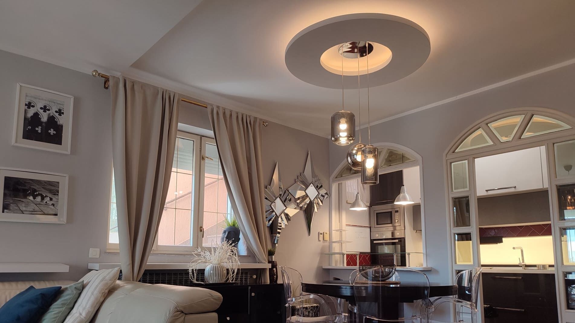 Uma sala de estar moderna com sofá, mesa de jantar com cadeiras, cozinha equipada e uma janela grande com cortinas.