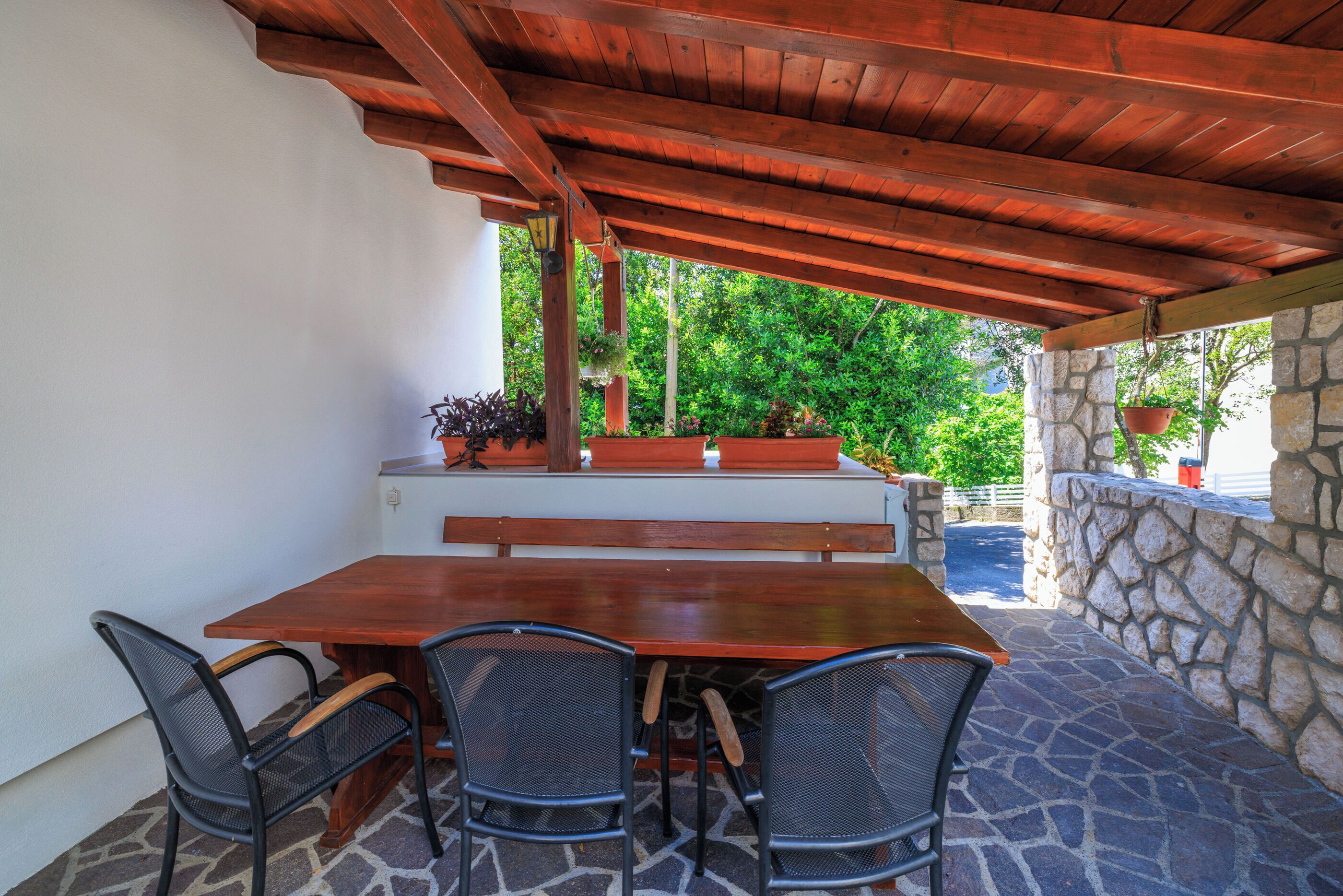 Um pátio externo com mesa e cadeiras de madeira, piso de pedra e uma parede de pedra.