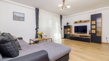 Uma sala de estar moderna com um sofá cinzento, piso de madeira, uma televisão e uma planta decorativa.