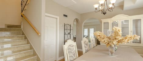 Uma sala de jantar com uma mesa comprida, cadeiras brancas, um candelabro e uma escadaria.