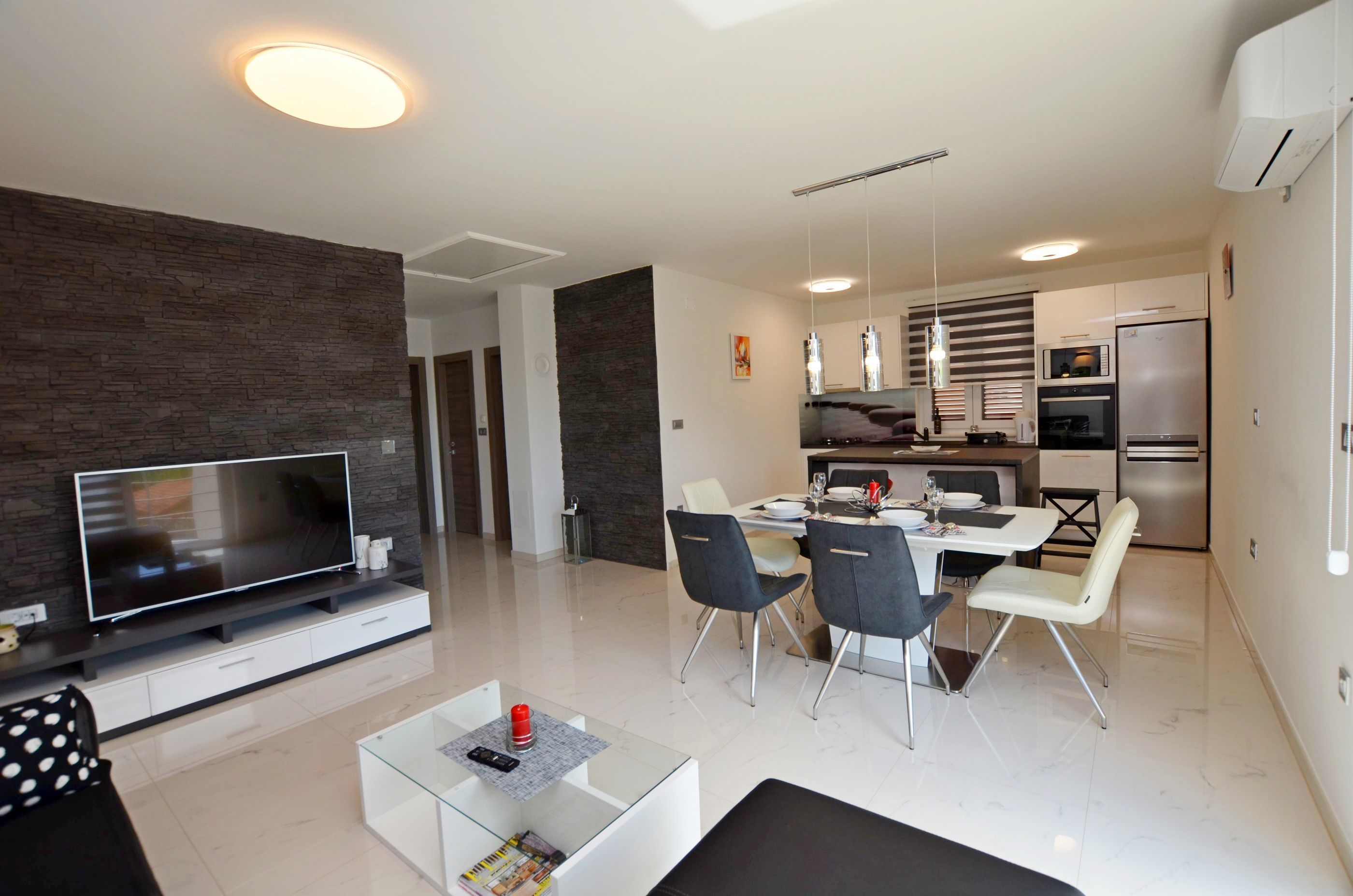 Uma sala de estar moderna com uma TV de tela plana, uma mesa de centro de vidro e uma área de jantar com mesa branca e cadeiras pretas.