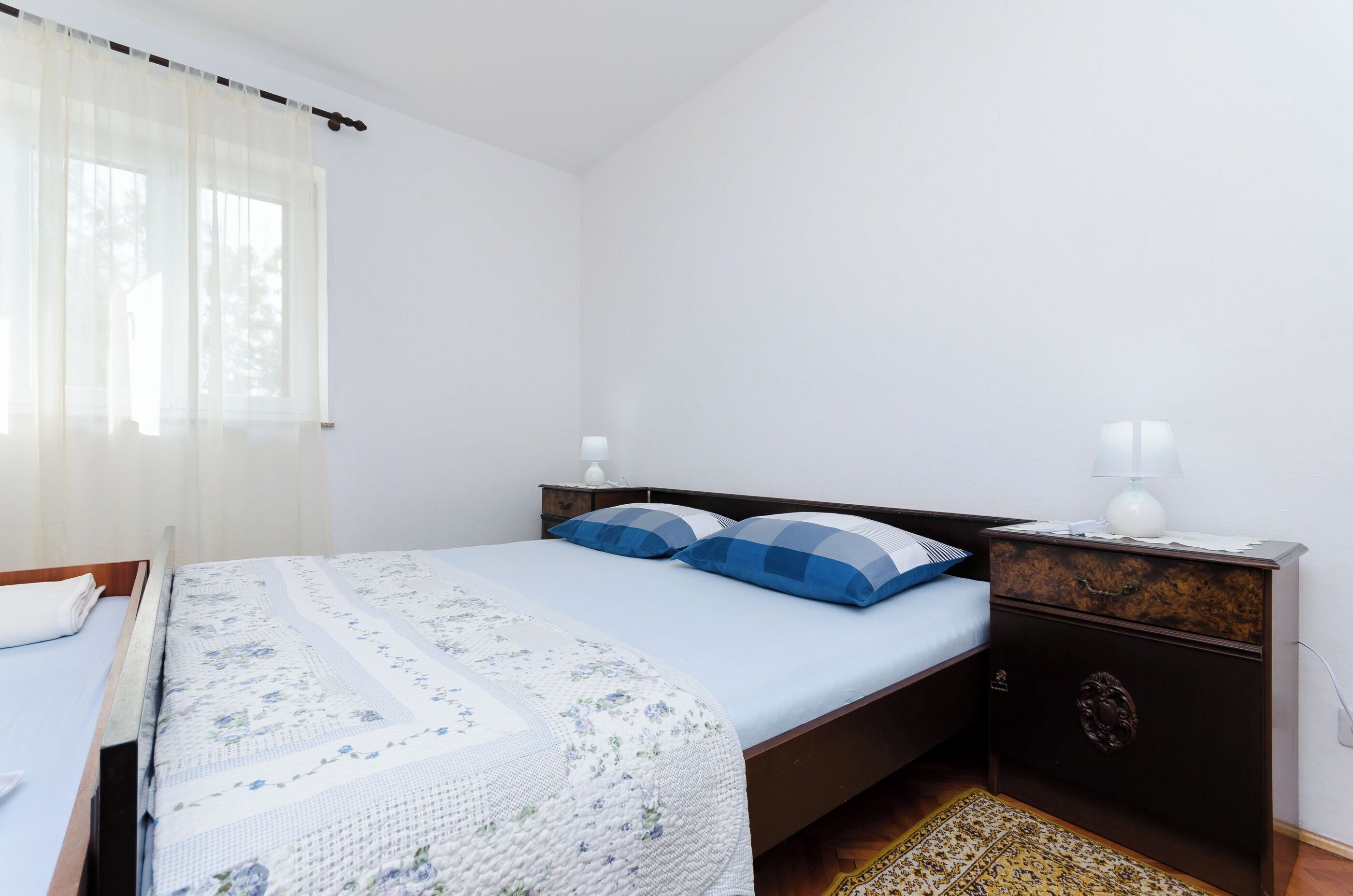 Uma cama bem arrumada com colcha floral e duas almofadas azuis, uma mesa de cabeceira com abajur, e um criado-mudo de madeira com gaveta.
