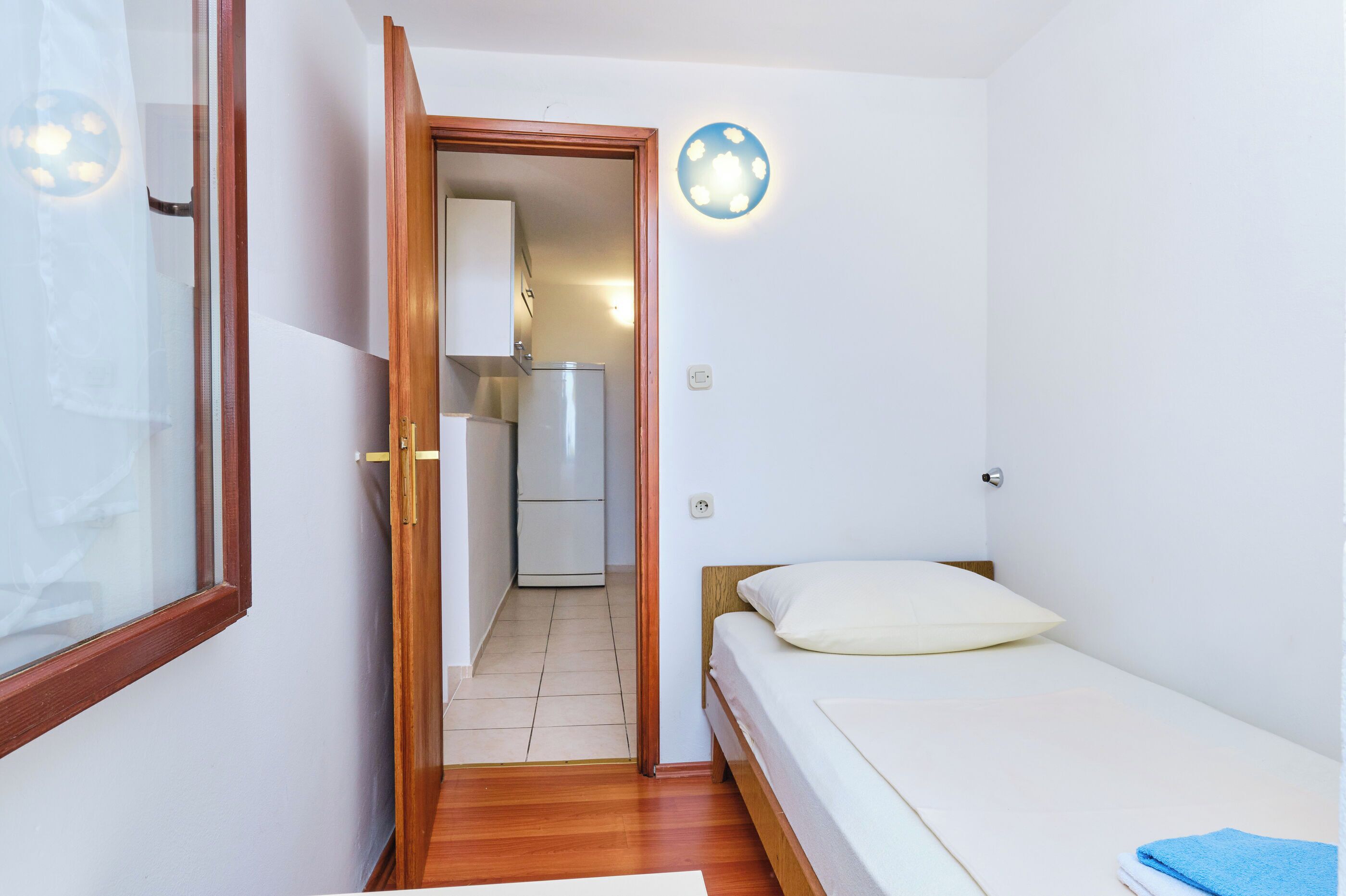Um quarto pequeno e bem iluminado, com uma cama, uma porta de madeira que dá para uma cozinha e uma luminária fixada na parede.