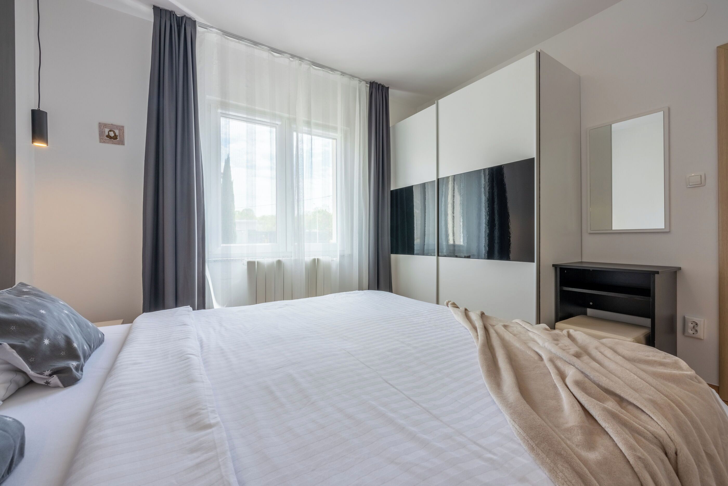 Quarto de hotel moderno com uma cama grande, uma mesinha de cabeceira, um espelho e uma janela com cortinas.
