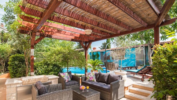 Uma área de estar coberta ao ar livre, com móveis de vime, piscina ao fundo e vista para um edifício com fachada azul.