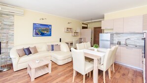 Uma sala moderna com sofá, mesa de centro e área de refeições. Há uma cozinha com armários e geladeira. Um quadro está na parede.