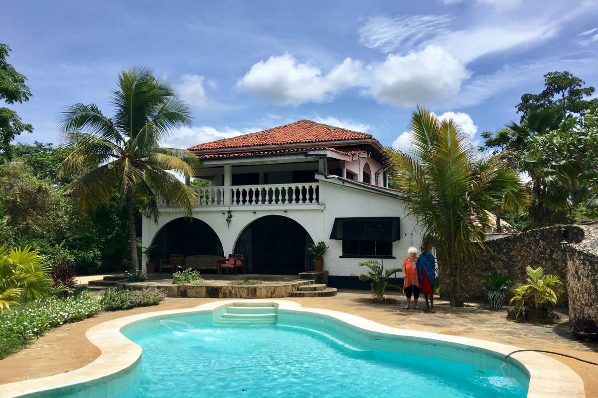 Uma área de piscina com borda curva, um edifício branco com telhado de telhas vermelhas e palmeiras.