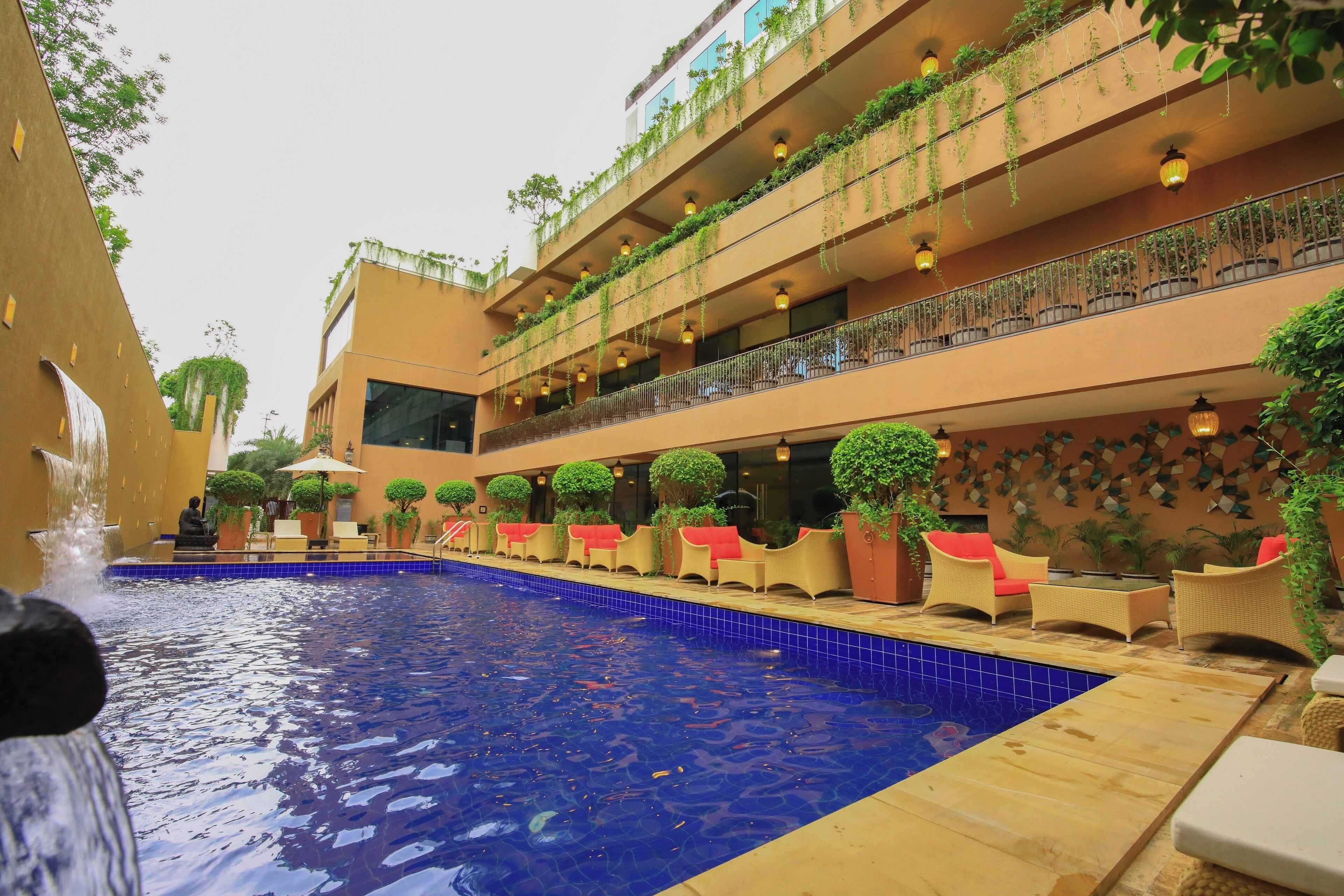 Piscina com espreguiçadeiras e mesas, cercada por um edifício com varandas e plantas.