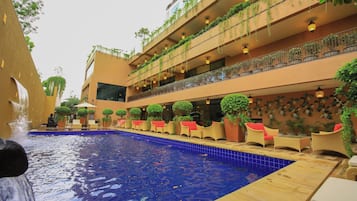 Piscina com espreguiçadeiras e mesas, cercada por um edifício com varandas e plantas.