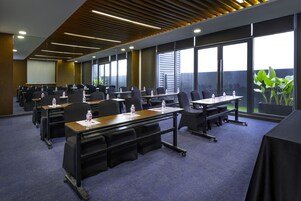 Uma sala de conferências com várias mesas e cadeiras, equipada com garrafas de água, e grandes janelas com vista para o verde lá fora.