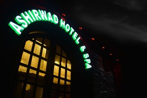 Exterior do Hotel & Spa Ashirwad com letreiros iluminados.