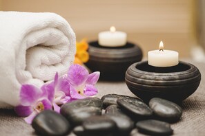 Ambiente de spa com toalha branca enrolada, duas velas acesas em castiçais pretos e flores roxas.