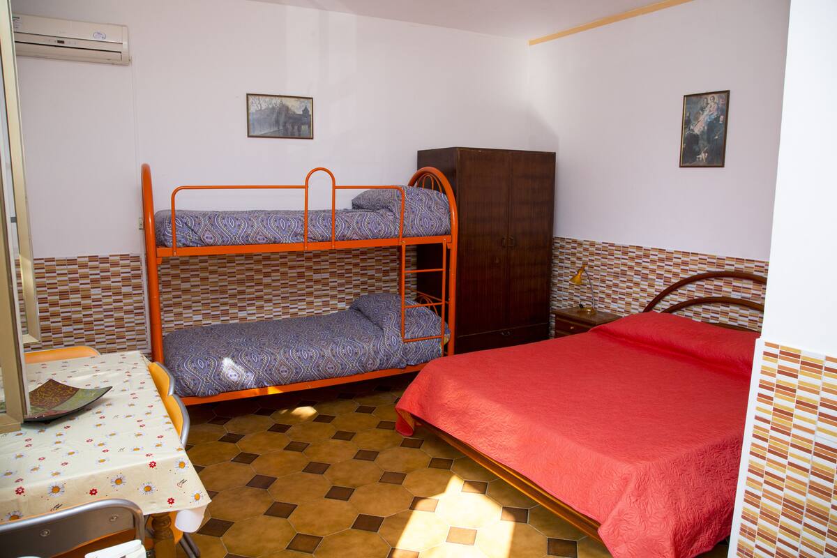 Quarto com beliches, guarda-roupa de madeira, mesa com toalha de mesa estampada e colcha vermelha.
