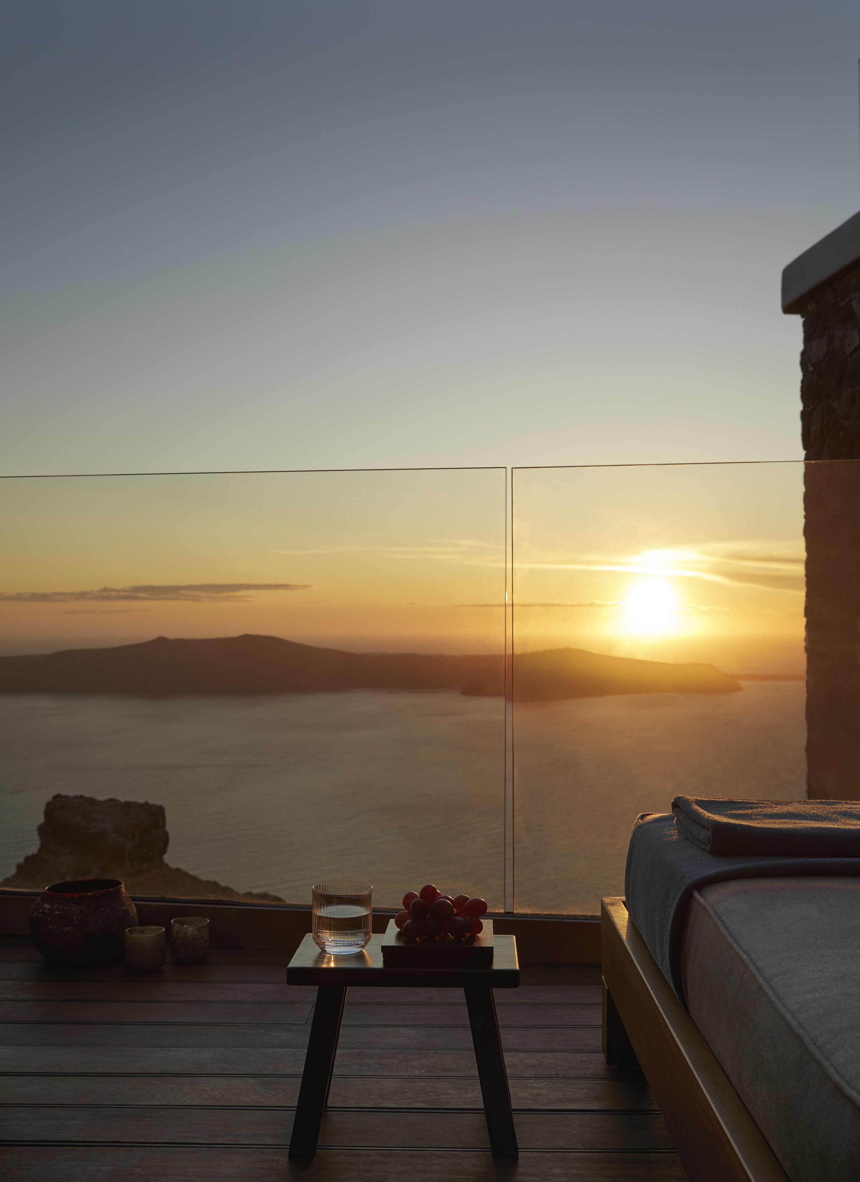 Uma varanda com vista para o pôr do sol sobre o mar, uma mesa com um copo de água e um vaso de flores, e uma cama com um cobertor azul.