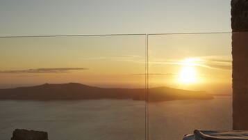 Uma varanda com vista para o pôr do sol sobre o mar, uma mesa com um copo de água e um vaso de flores, e uma cama com um cobertor azul.
