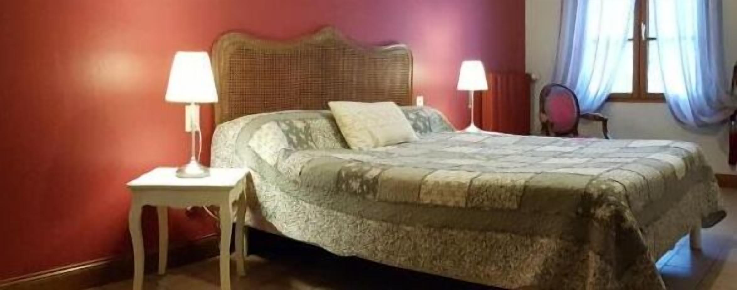 Quarto com cama, duas luminárias de cabeceira, janela com cortinas e uma mesinha.