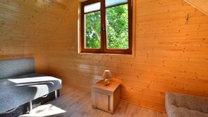 Quarto de cabana de madeira com uma cama, uma pequena mesa de cabeceira com abajur e uma janela com vista para árvores.