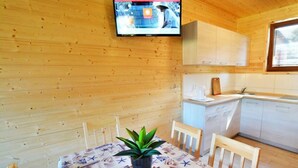 Interior de uma cabana de madeira com uma mesa de jantar, cadeiras, um vaso de planta, uma televisão presa à parede e uma área de cozinha.