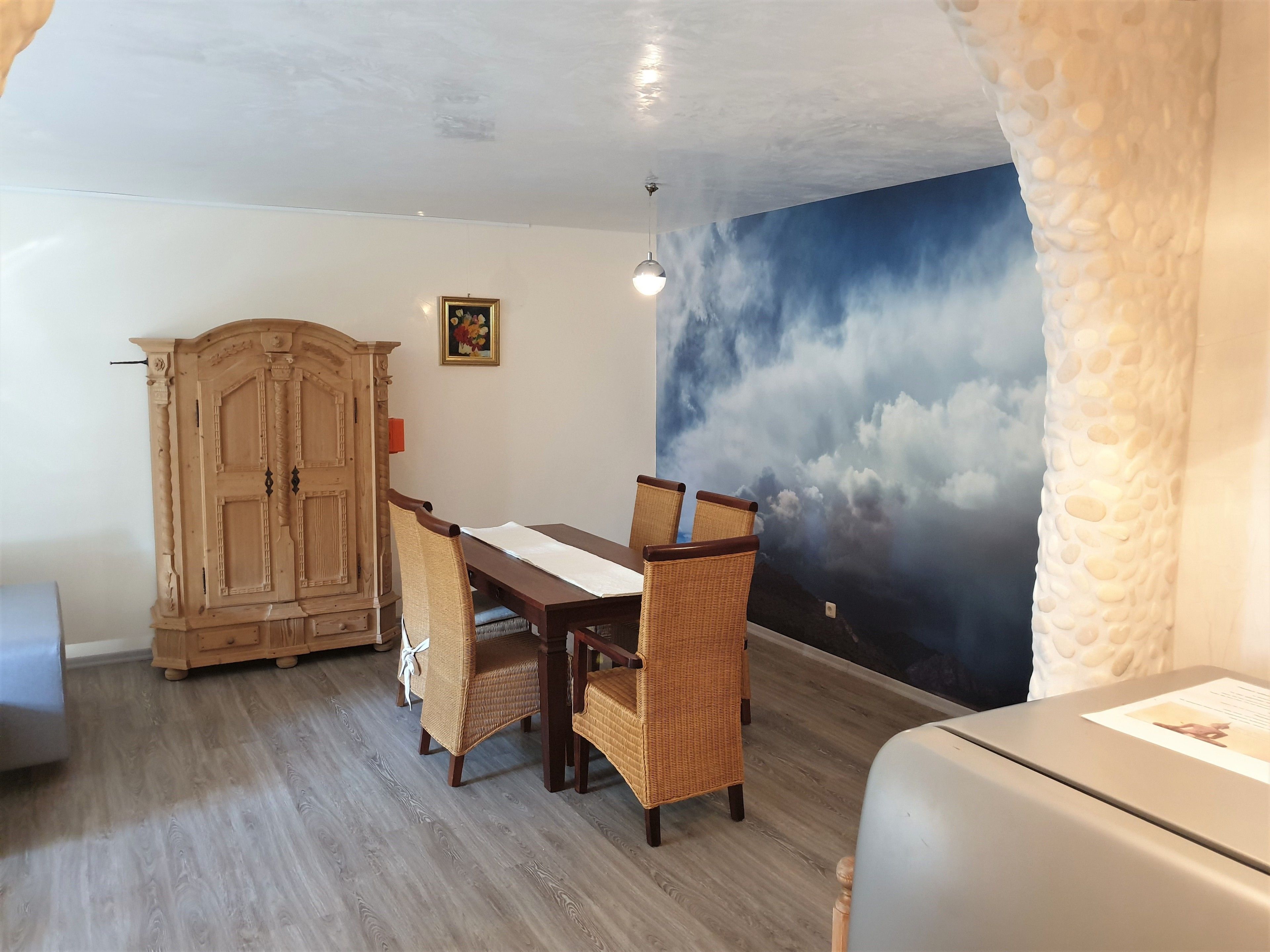 Uma área de refeições com mesa de madeira e cadeiras de vime, um armário de madeira e um mural na parede com tema de nuvens.