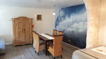 Uma área de refeições com mesa de madeira e cadeiras de vime, um armário de madeira e um mural na parede com tema de nuvens.