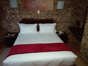 Quarto de hotel com parede de tijolos, cama com roupa de cama branca e manta vermelha, duas mesas de cabeceira com abajures e um telefone em uma das mesas.