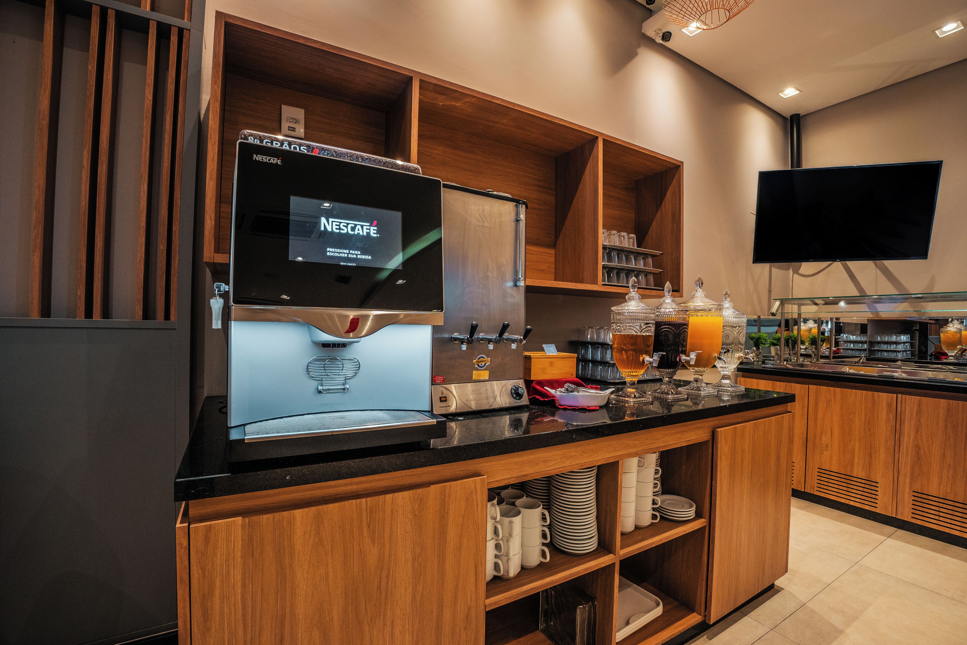 Uma estação de café moderna com máquina Nescafé, televisão de tela plana e diversas bebidas.
