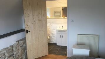 Quarto com porta de madeira, parede de pedra, cama com cobertor, mesa de cabeceira branca e banheiro com pia e armários.