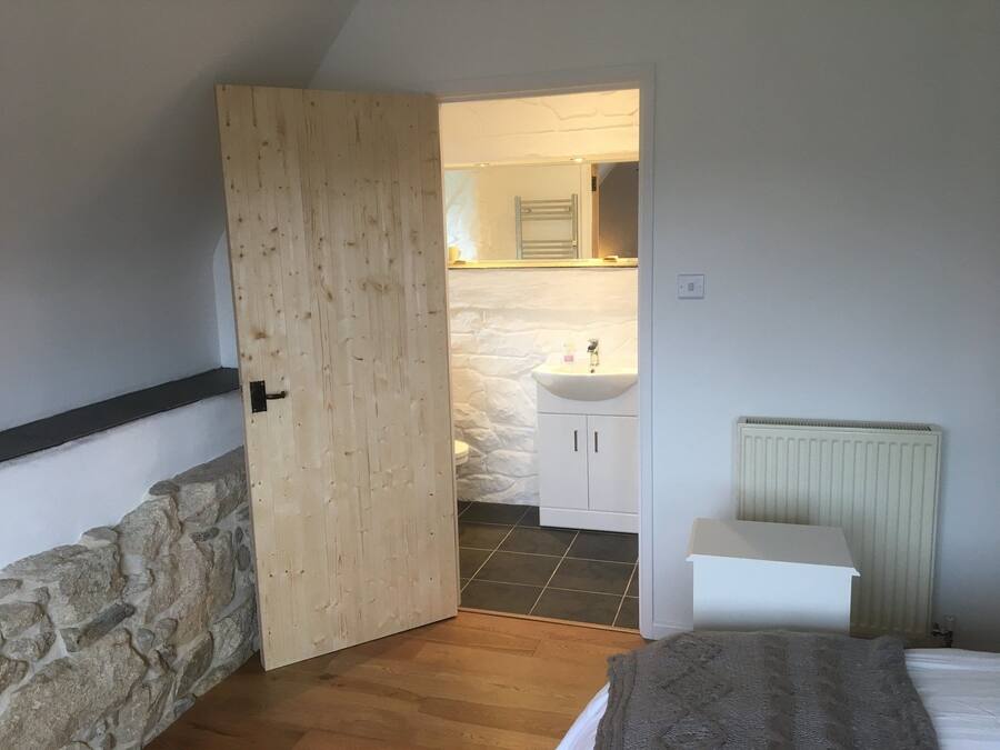 Quarto com porta de madeira, parede de pedra, cama com cobertor, mesa de cabeceira branca e banheiro com pia e armários.