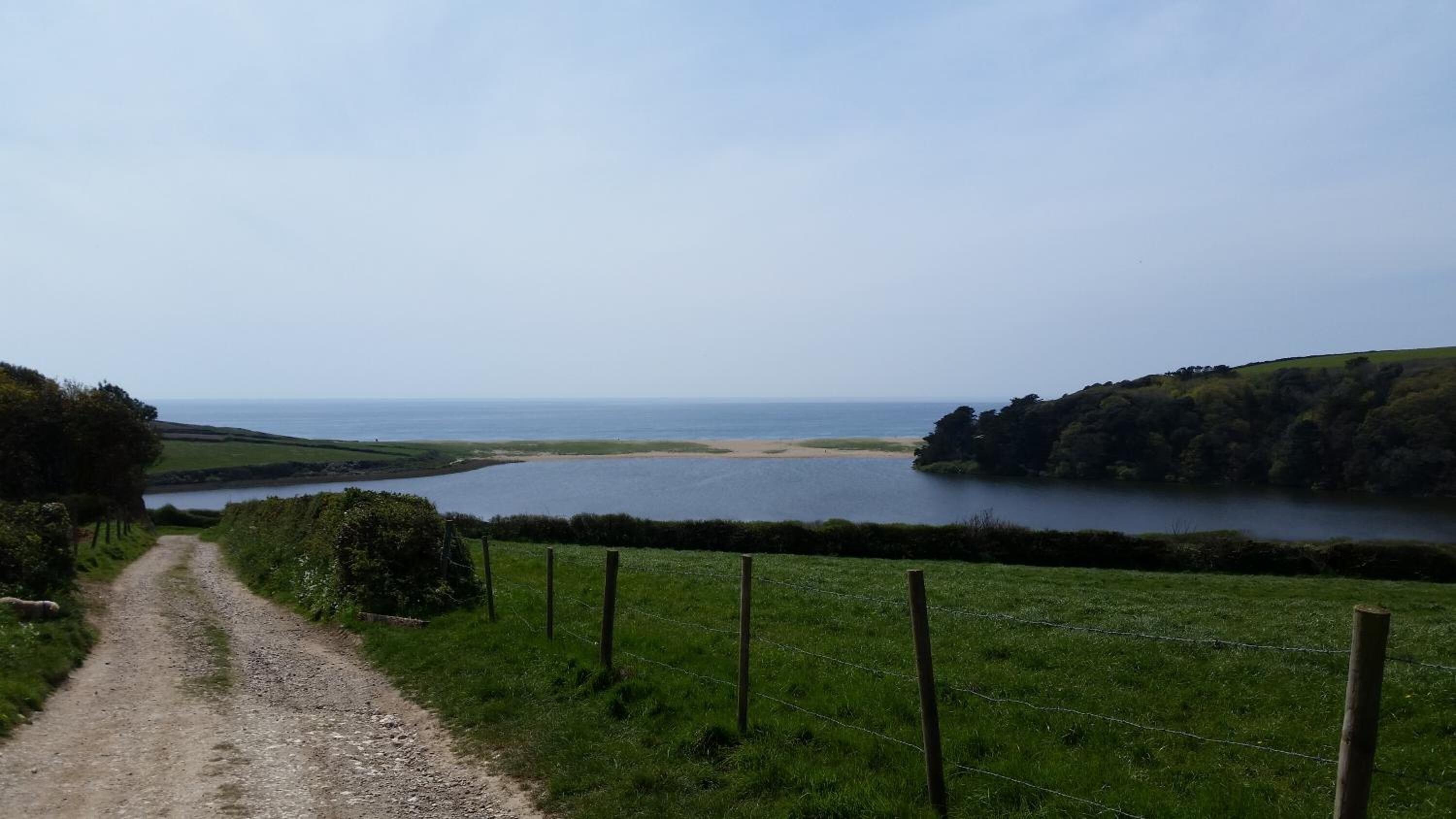 Uma trilha de cascalho leva a uma área gramada cercada, com vista para um lago e colinas ao longe.