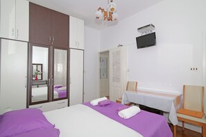 Quarto de hotel com cama, guarda-roupa, escrivaninha e cadeira, e televisão na parede.