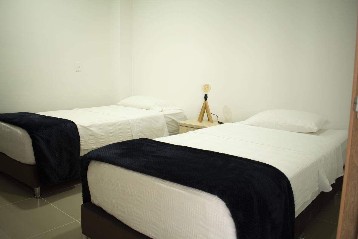 Quarto com duas camas de solteiro, cada uma com colchão branco e manta azul escura. Há uma mesa de cabeceira com abajur entre as camas.