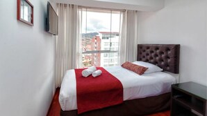 Quarto de hotel com uma cama grande, manta vermelha e vista para edifícios pela janela.