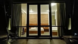 Quarto de hotel com uma grande porta de vidro deslizante que dá para uma varanda, uma cama com roupa de cama branca e uma cabeceira de madeira.