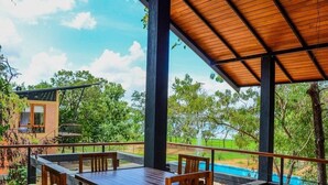 Área externa coberta com piscina, móveis de madeira e vista para um edifício e árvores.