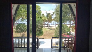 Vista de um quarto com porta de vidro que dá para área externa com praia, palmeiras e deck de madeira com cadeiras.