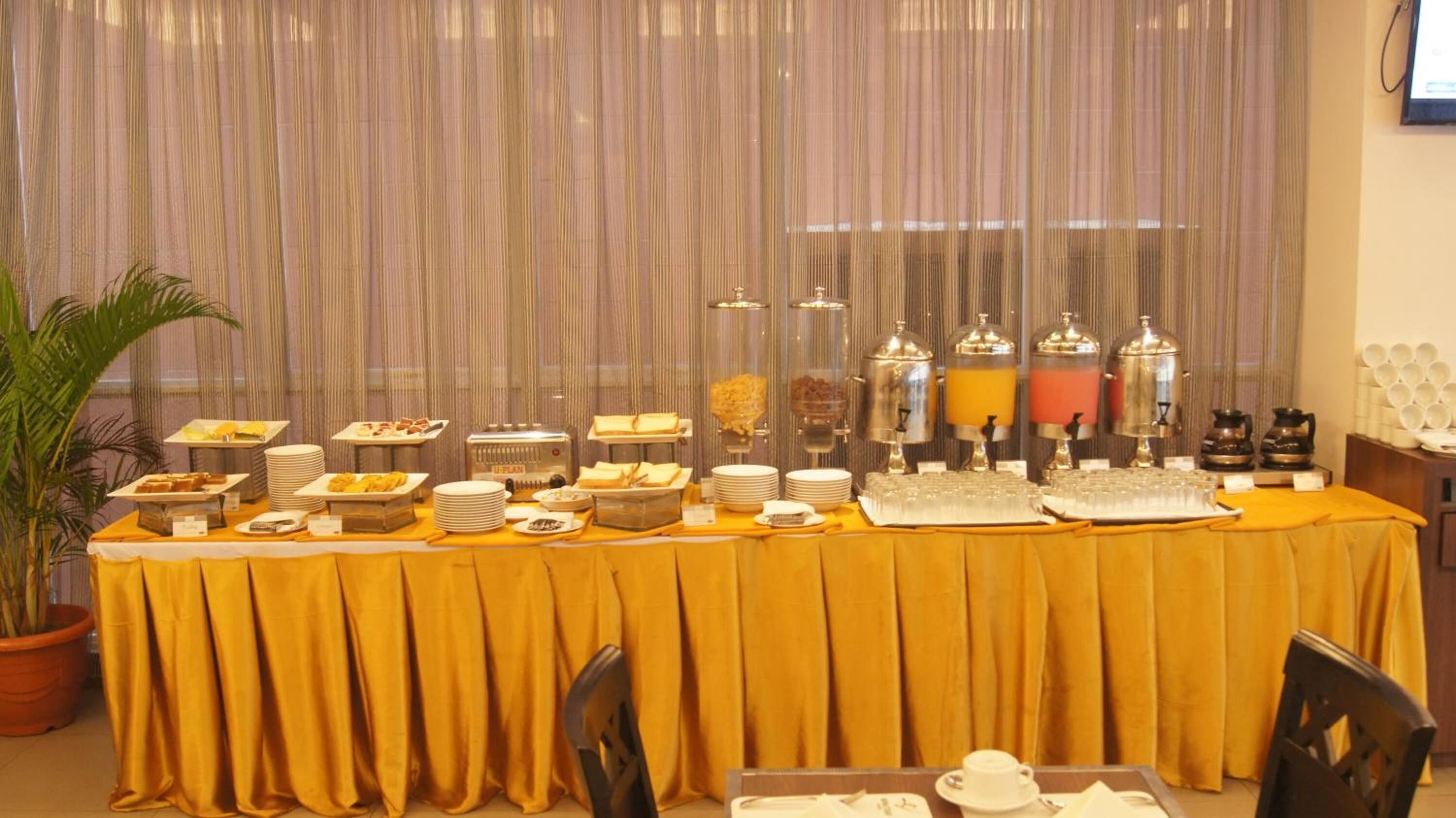 Mesa de buffet com diversos alimentos e dispensadores de bebidas, com cortinas leves ao fundo.
