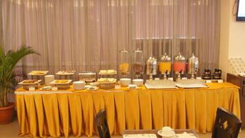 Mesa de buffet com diversos alimentos e dispensadores de bebidas, com cortinas leves ao fundo.