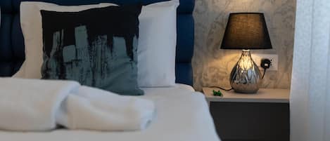 Cama bem arrumada com lençóis brancos, cabeceira azul e almofada decorativa. Há um abajur em uma mesa de cabeceira ao lado da cama.