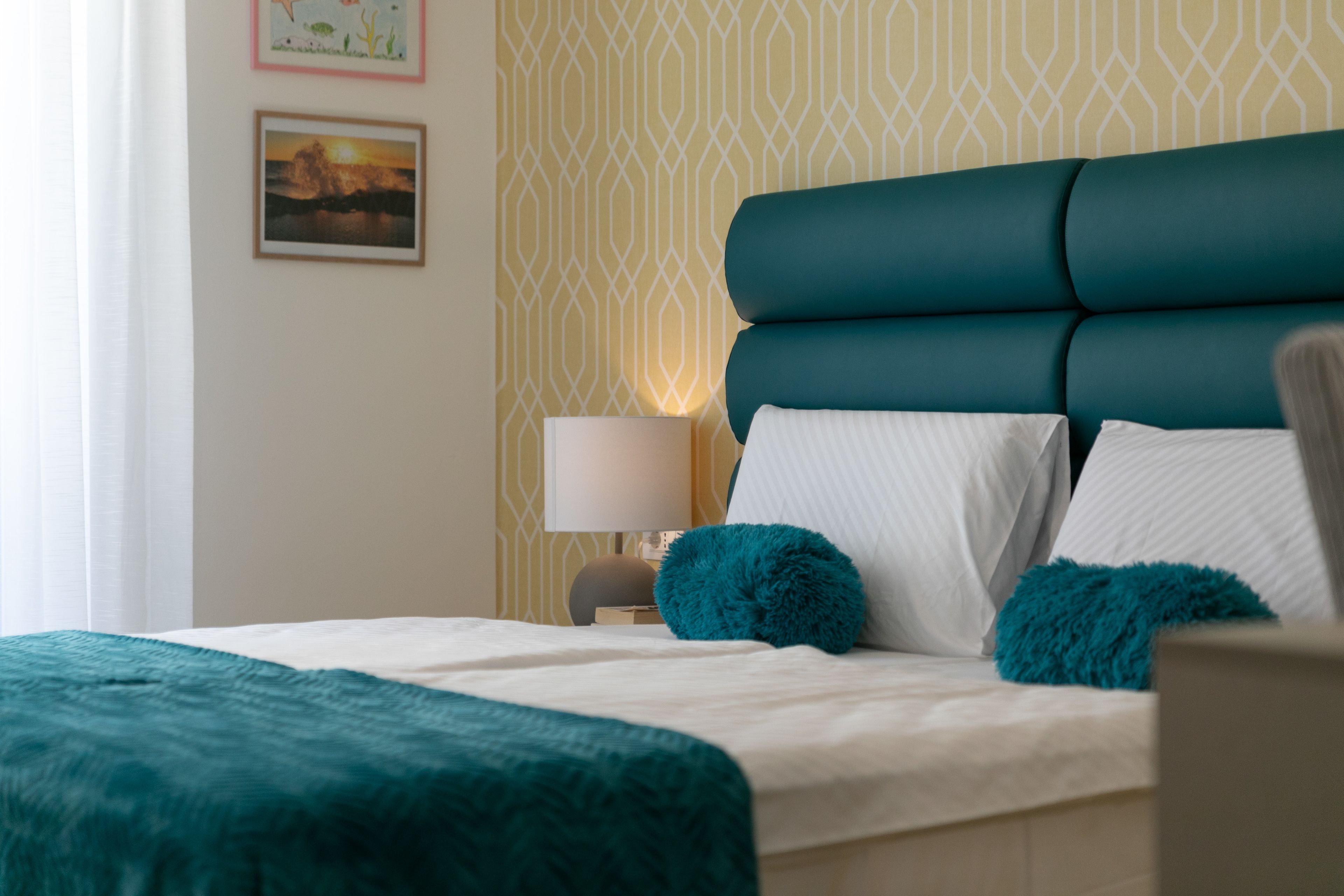 Uma cama bem arrumada com roupa de cama e almofadas verde-água, um abajur e uma parede com padrão geométrico.