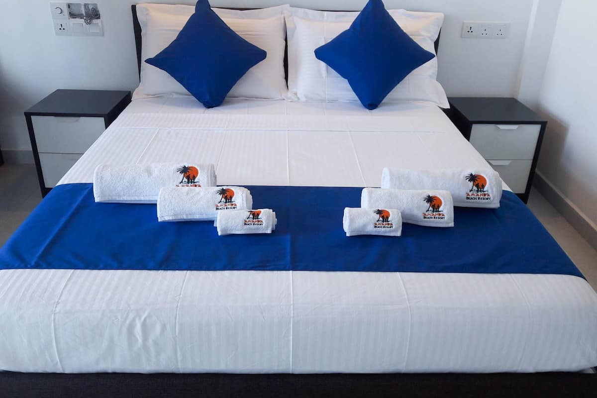 Cama bem arrumada com lençóis brancos, dois travesseiros azuis e um cobre-leito azul. Quatro toalhas com o logotipo de uma palmeira.