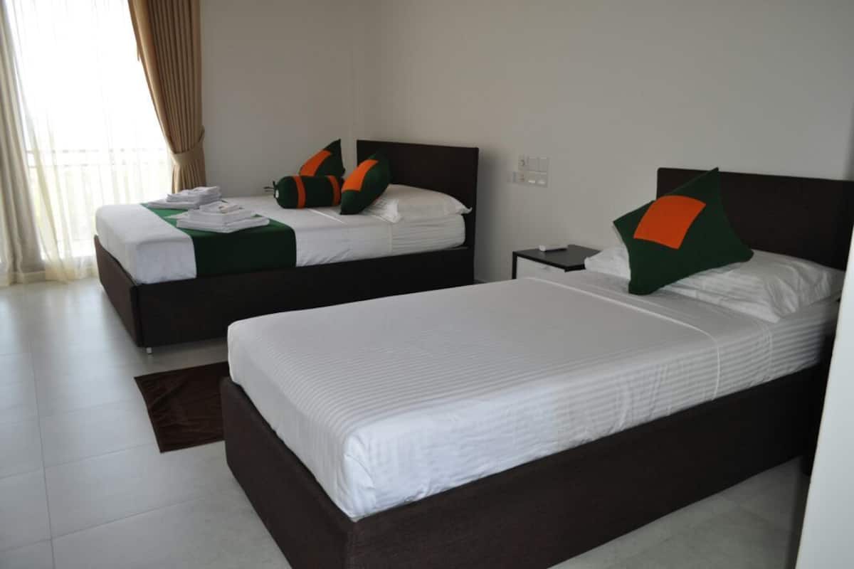 Quarto de hotel com duas camas, cada uma com lençóis brancos e almofadas verdes e laranja. Há uma pequena mesa de cabeceira com uma luminária de um lado e uma tomada elétrica do outro.