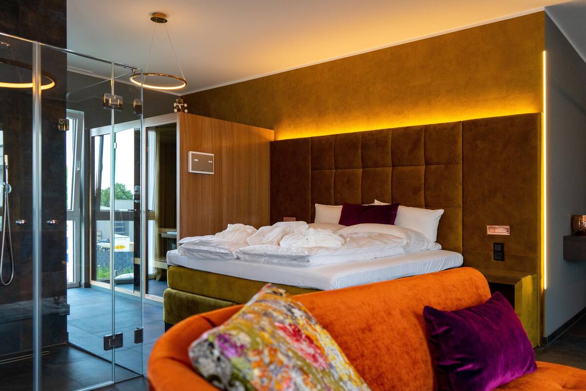Quarto de hotel moderno com uma cama grande, box de vidro para o chuveiro e vista para o exterior.