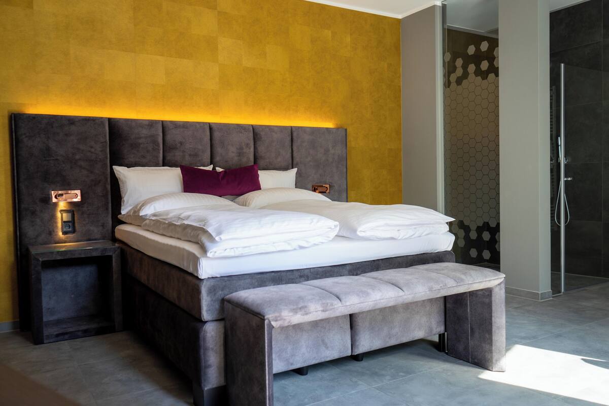 Quarto moderno com uma cama grande, uma mesa de cabeceira com abajur e um banco aos pés da cama.