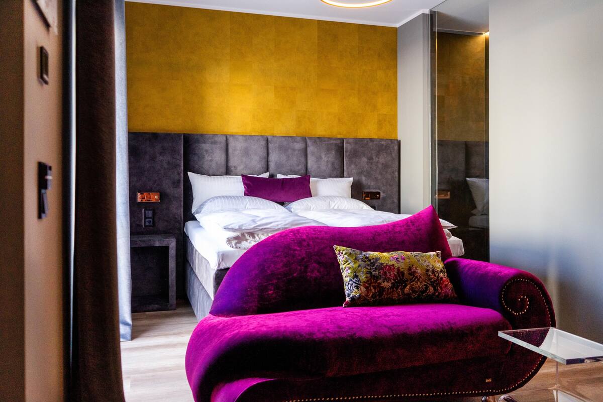 Quarto de hotel moderno com uma cama grande, um sofá roxo e uma parede amarela de destaque.