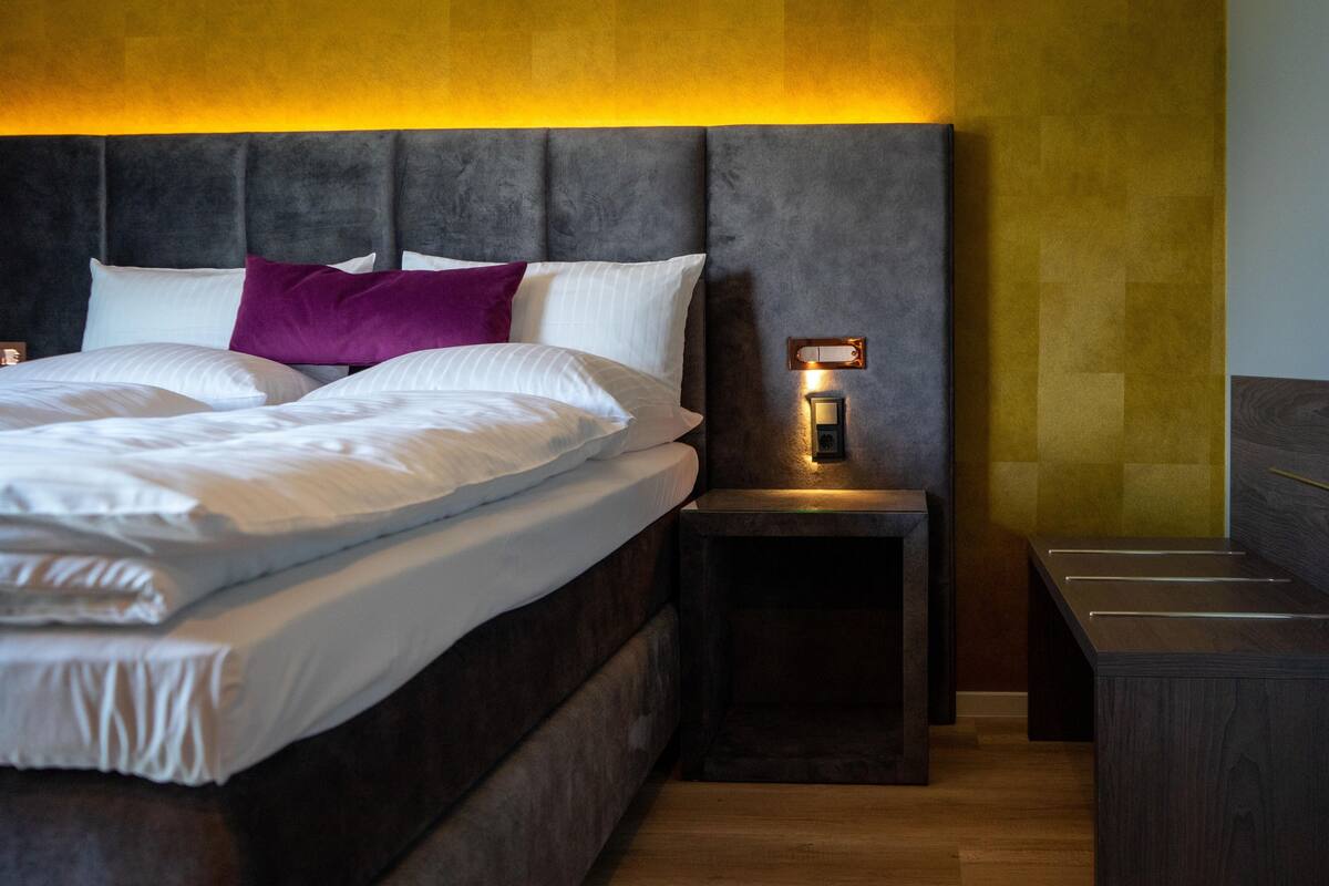 Quarto moderno com cama grande, cabeceira, mesas de cabeceira e luminária fixada na parede.