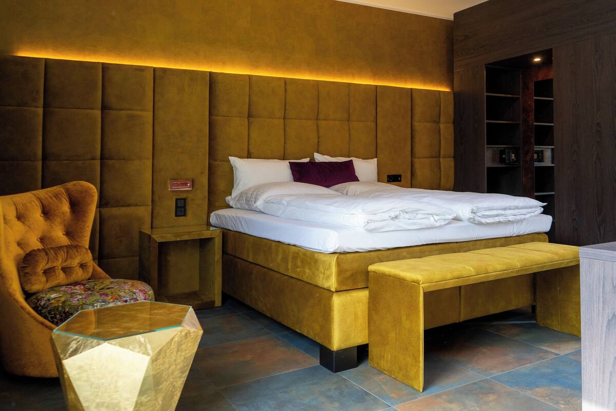 Quarto moderno com uma cama grande, um banco, uma cadeira e uma mesinha de cabeceira.