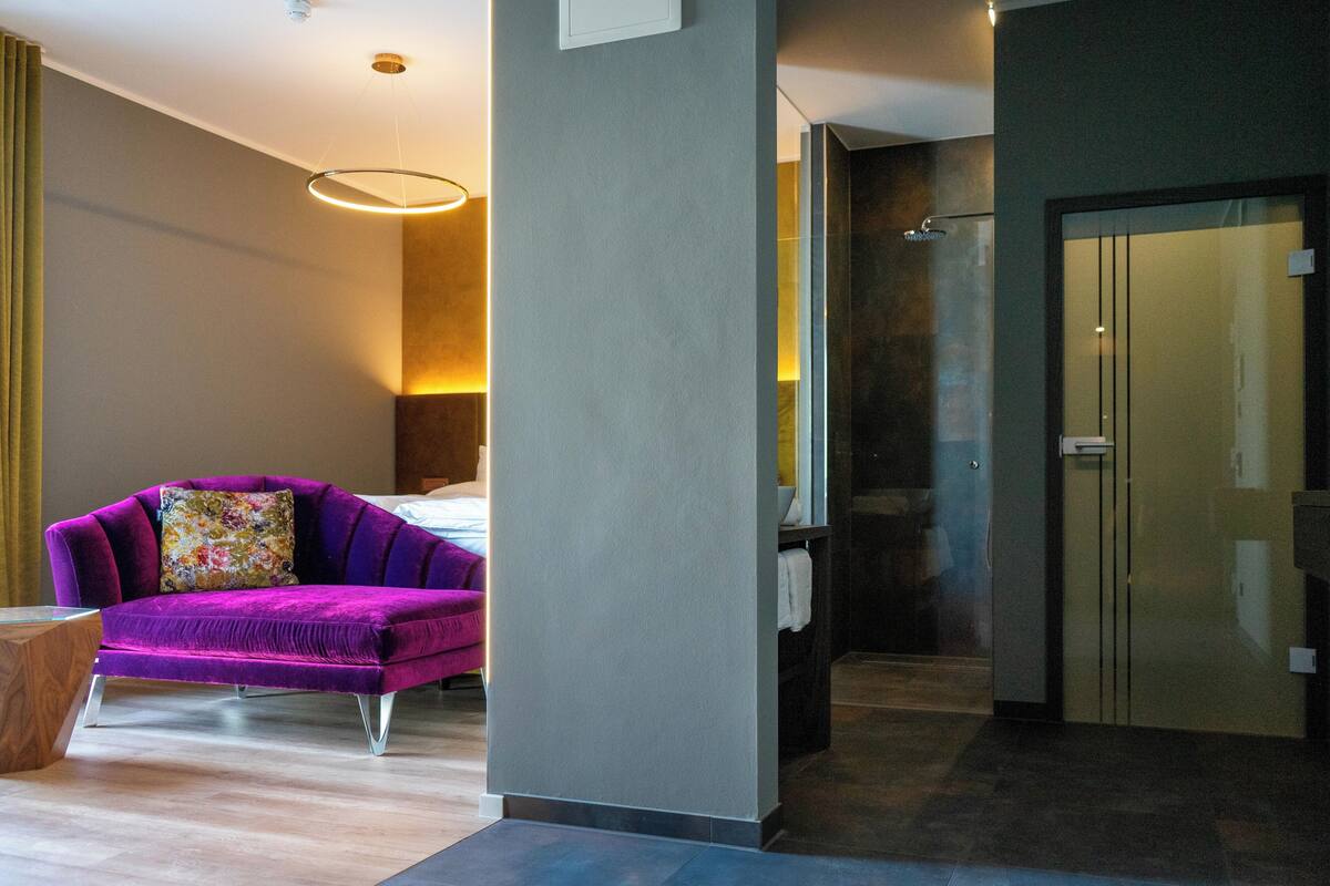 Quarto de hotel moderno com sofá roxo, mesa de apoio de madeira, área de chuveiro e guarda-roupa.