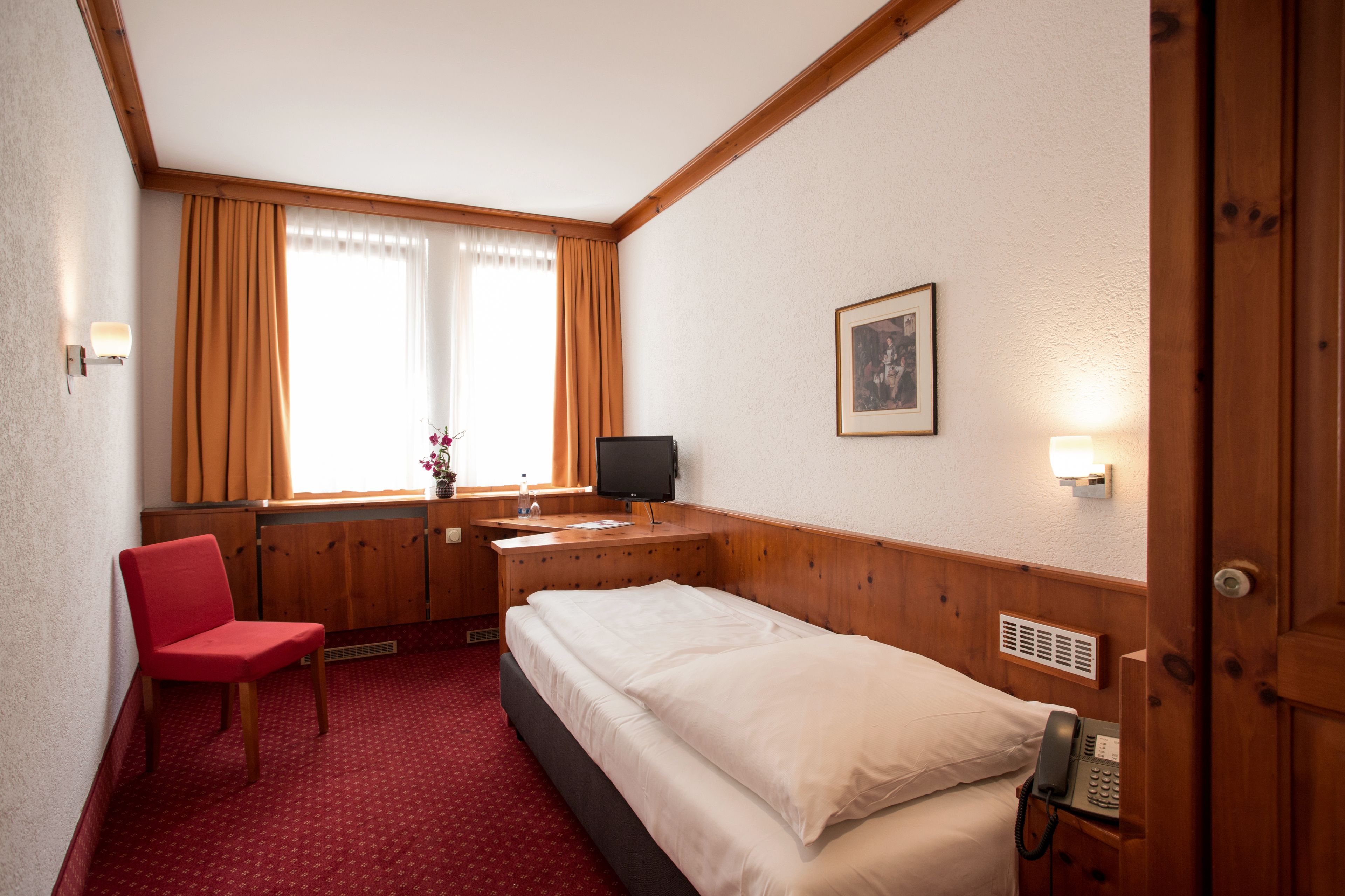 Quarto de hotel com cama, escrivaninha com computador, cadeira, telefone e janela com cortinas.